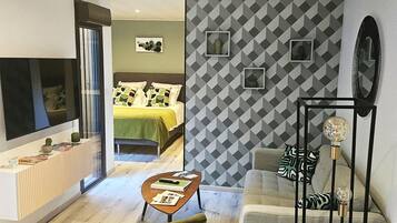 Uma sala de estar moderna com sofá, poltrona e mesa de centro. Há uma cama com colcha verde ao fundo.