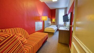 Quarto de hotel com parede vermelha, sofá listrado, cama, escrivaninha com luminária e televisão.