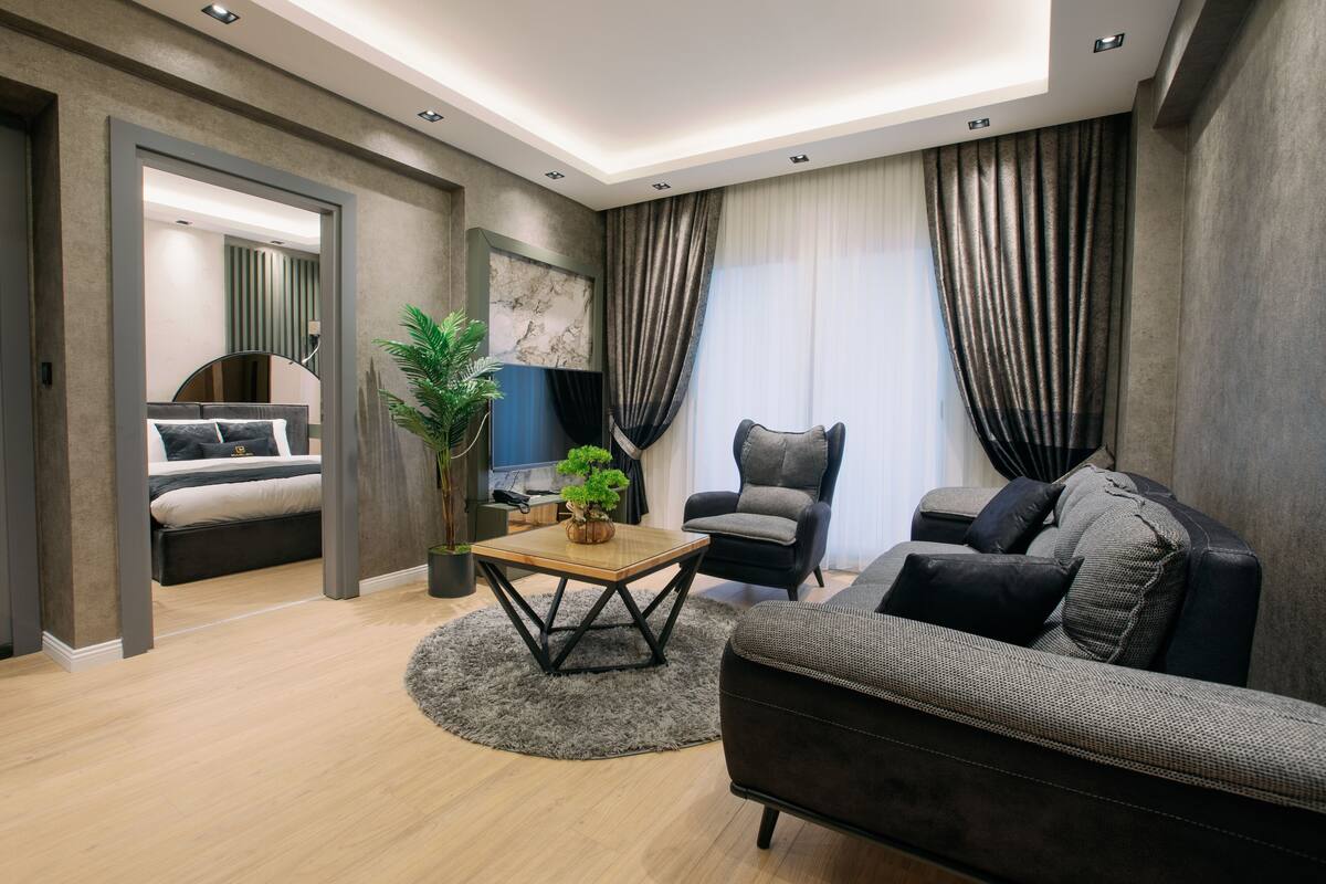 Uma sala de estar moderna com um sofá, poltrona e uma pequena mesa de centro de madeira. Há uma cama ao fundo e um vaso com planta sobre a mesa.