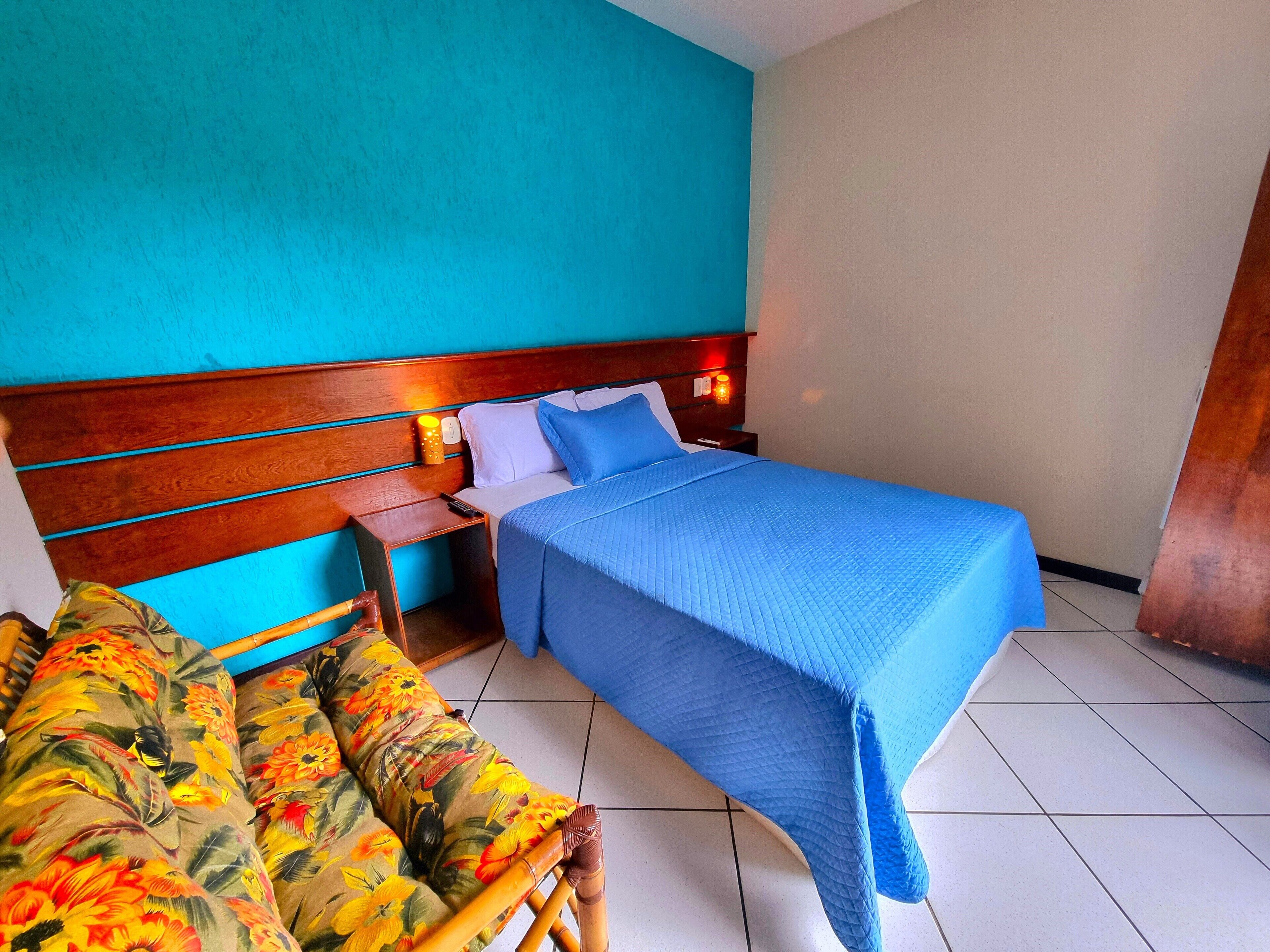 Quarto de hotel com uma cama, mesa de cabeceira, cabeceira de madeira, colcha azul, sofá floral e piso de azulejos.
