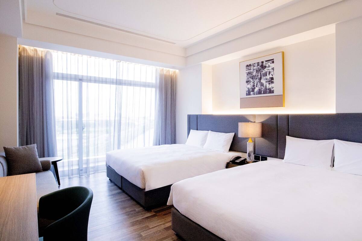 Quarto de hotel com duas camas, uma escrivaninha e uma cadeira. Há uma janela com cortinas, um quadro na parede e um abajur na mesa de cabeceira.