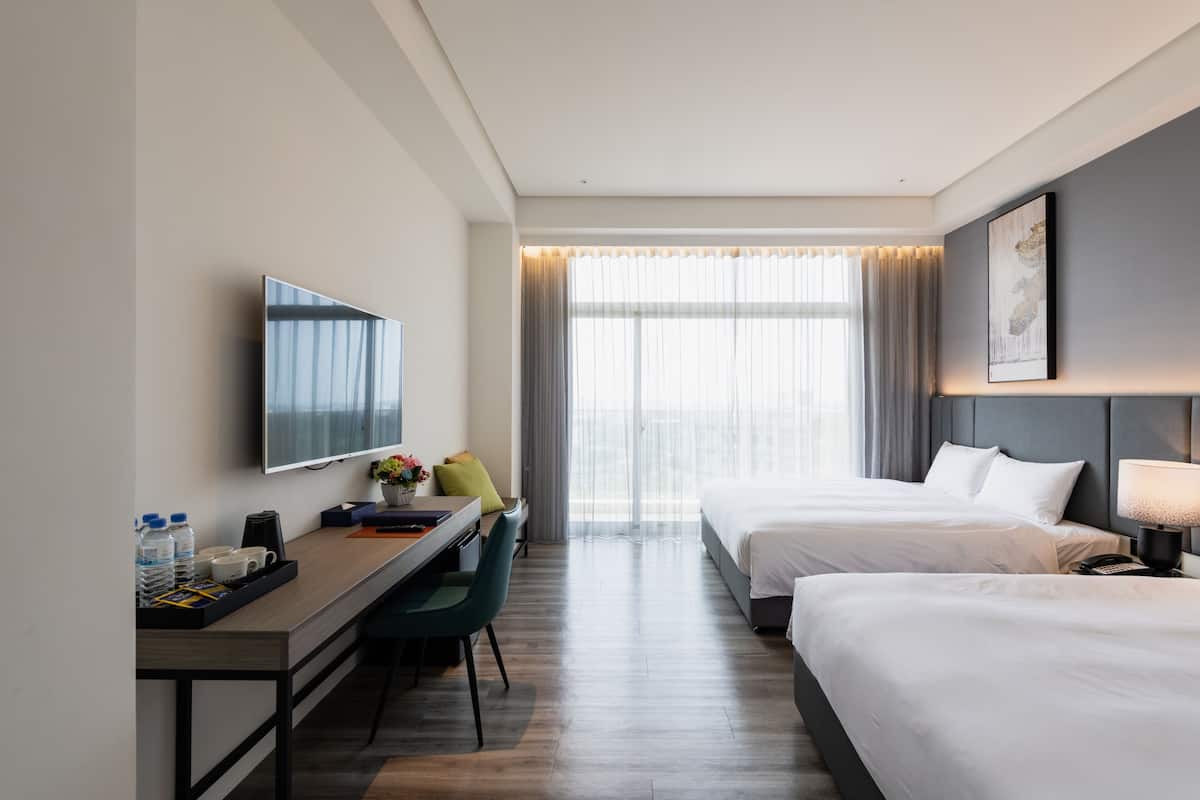 Quarto de hotel com duas camas, uma escrivaninha com cadeira, TV de tela plana e uma janela grande com cortinas.