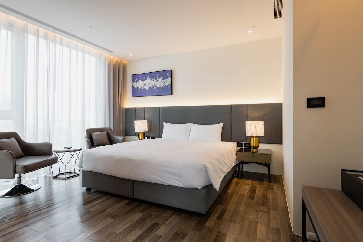 Quarto de hotel moderno com uma cama grande, duas mesas de cabeceira com abajures, um sofá e uma janela ampla com cortinas leves.