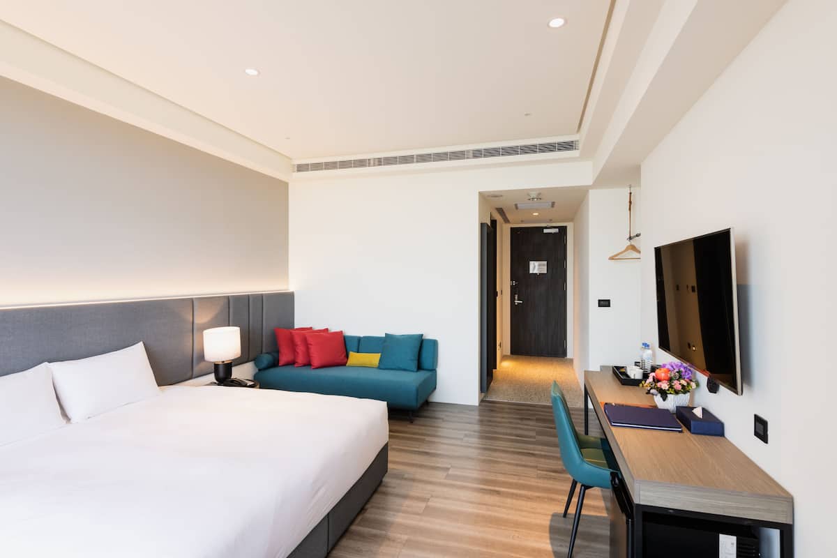 Quarto de hotel com cama, escrivaninha, sofá, televisão e porta que dá para outro cômodo.