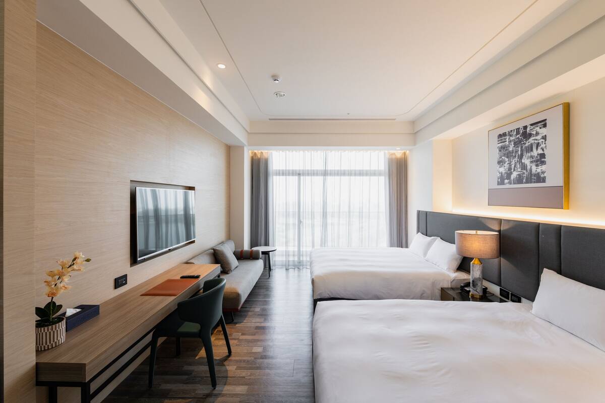 Quarto de hotel com duas camas, uma escrivaninha com cadeira, televisão e janela com cortinas.
