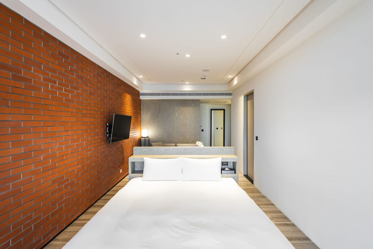 Quarto de hotel moderno com parede de tijolos, uma cama grande, televisão de tela plana, espelho e piso de madeira.