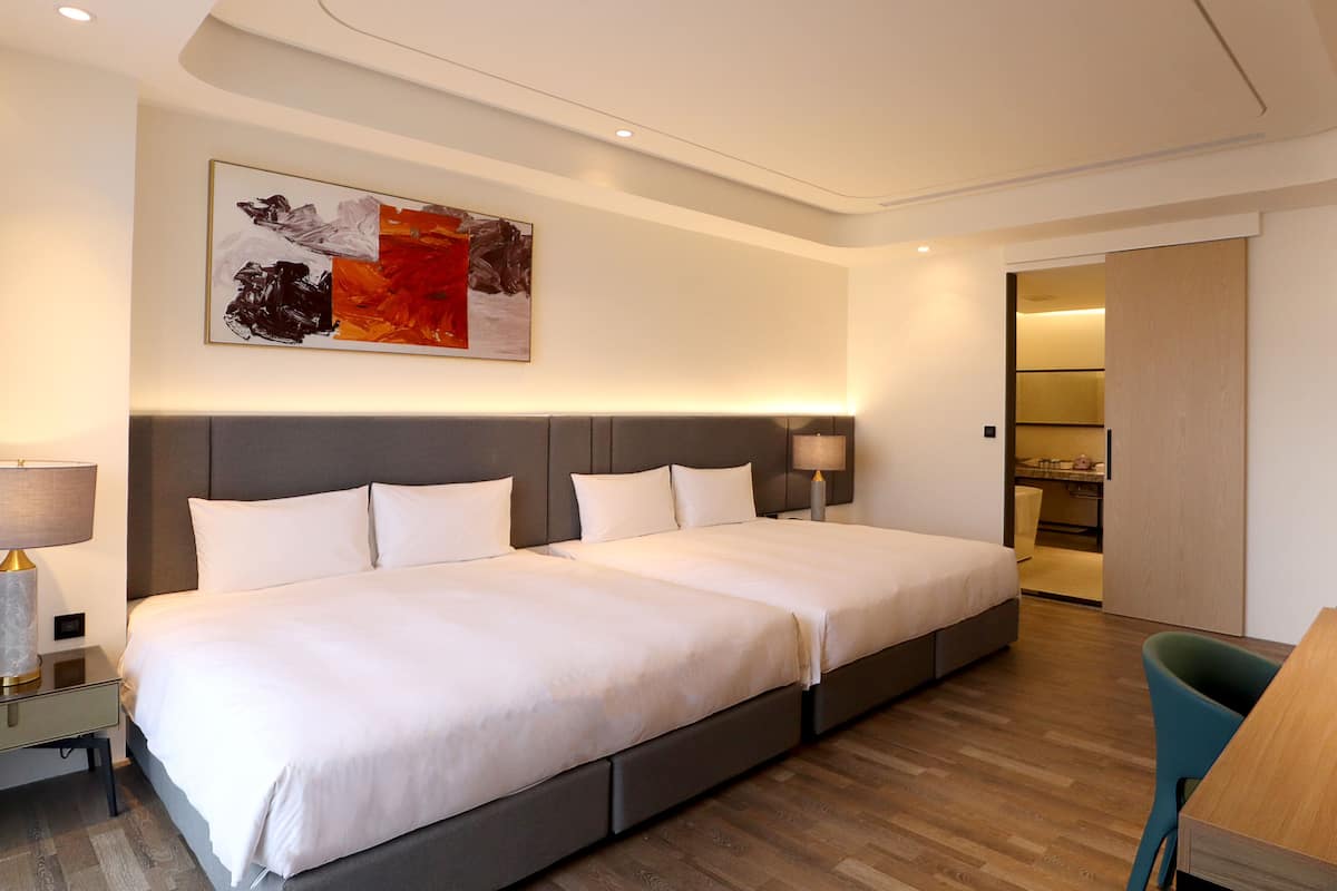 Quarto de hotel moderno com uma cama grande, abajures, piso de madeira e um quadro na parede.