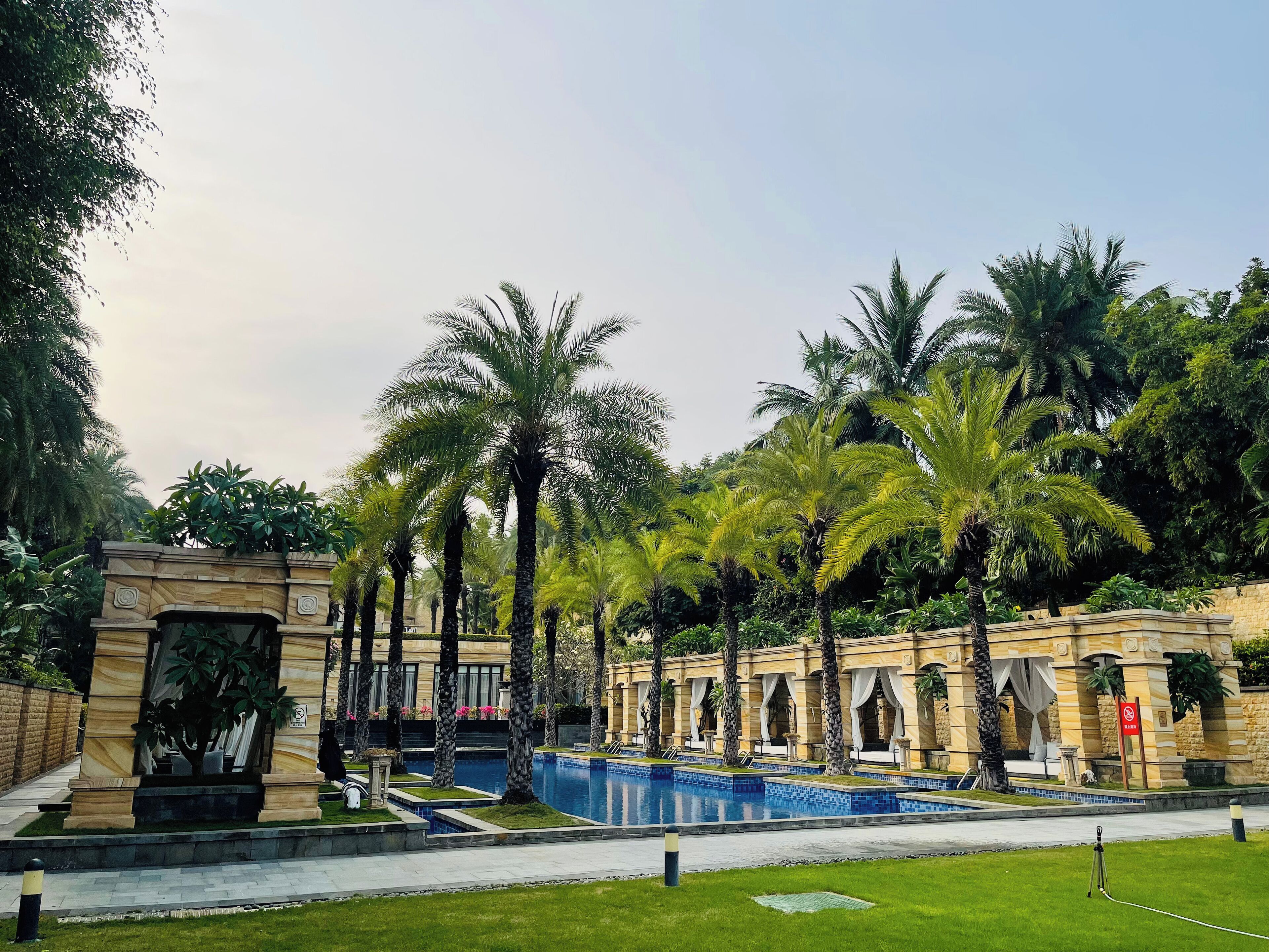 Uma área de piscina com palmeiras, um edifício de pedra e uma pequena estátua.