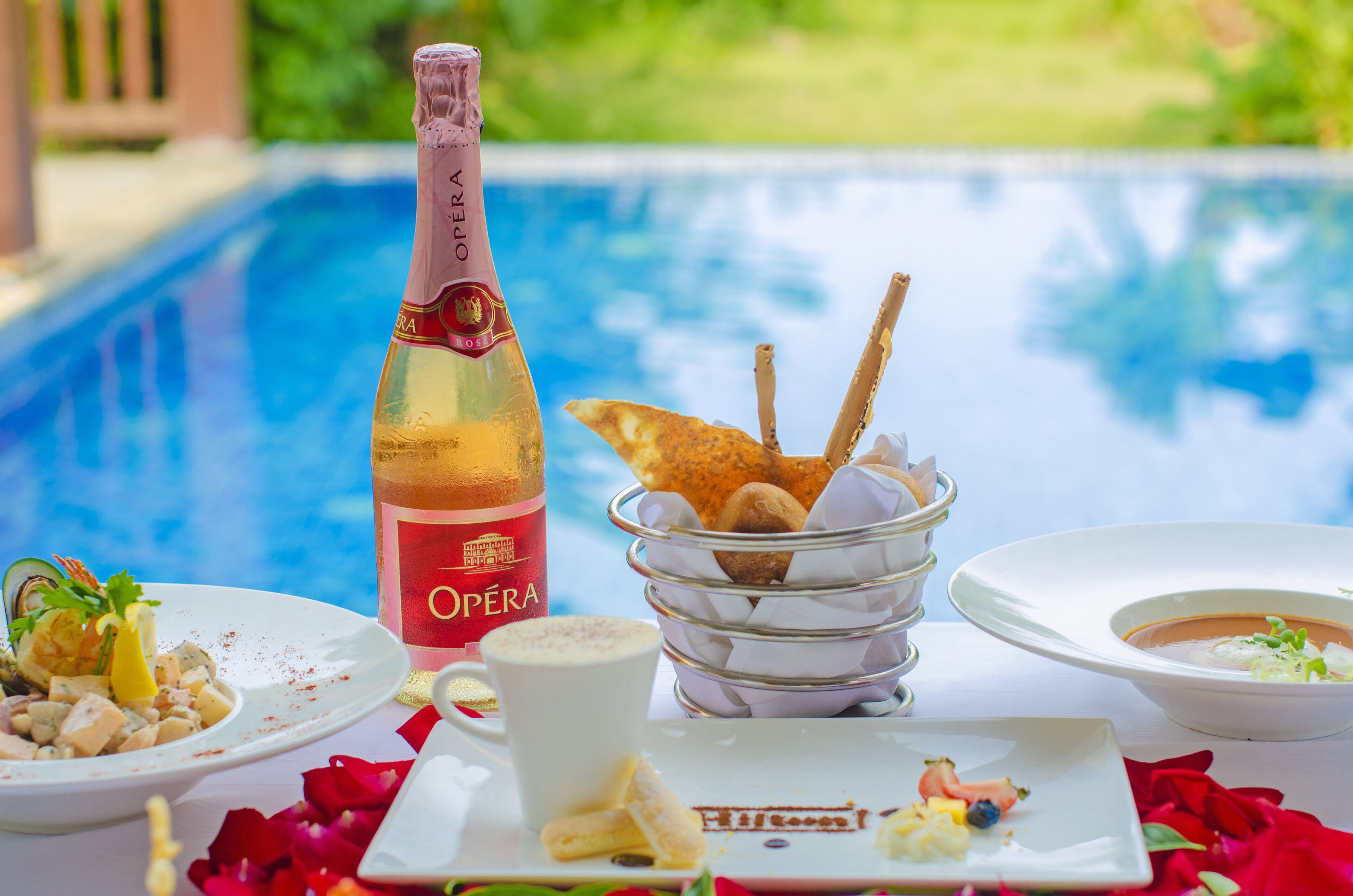 Disposição de refeições junto a uma piscina, com uma garrafa de champanhe, uma tigela de sopa e pratos de comida.