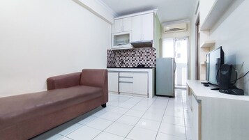 Uma cozinha compacta com armários brancos, micro-ondas e painel de azulejos. Há uma geladeira pequena e uma TV sobre um suporte.