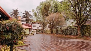 Rua residencial com casas em ambos os lados, pavimentada com tijolos vermelhos, cercada por vegetação e árvores.