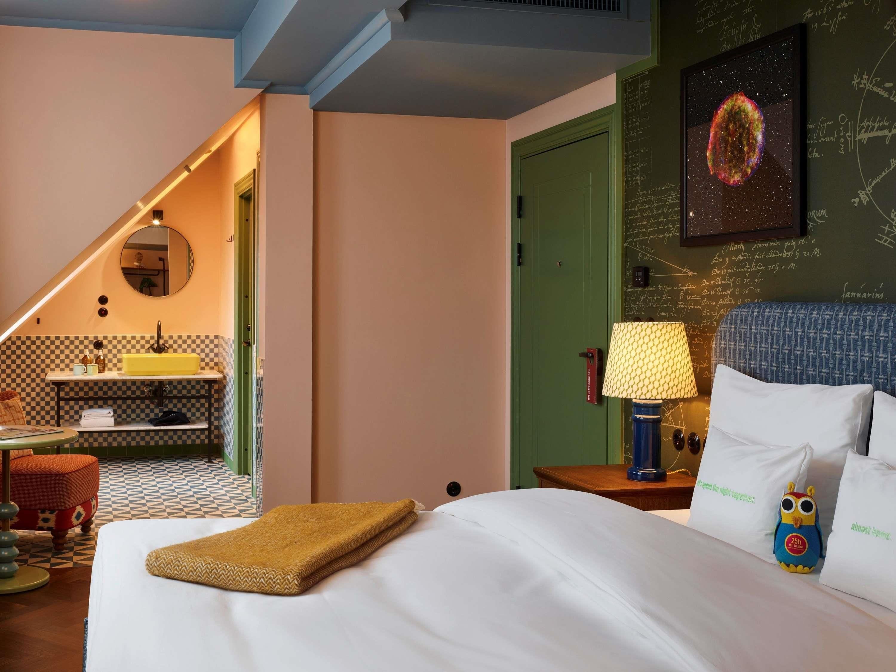 Quarto de hotel com uma cama, uma escrivaninha, uma luminária, uma parede com uma obra de arte de tema celestial e uma porta verde.