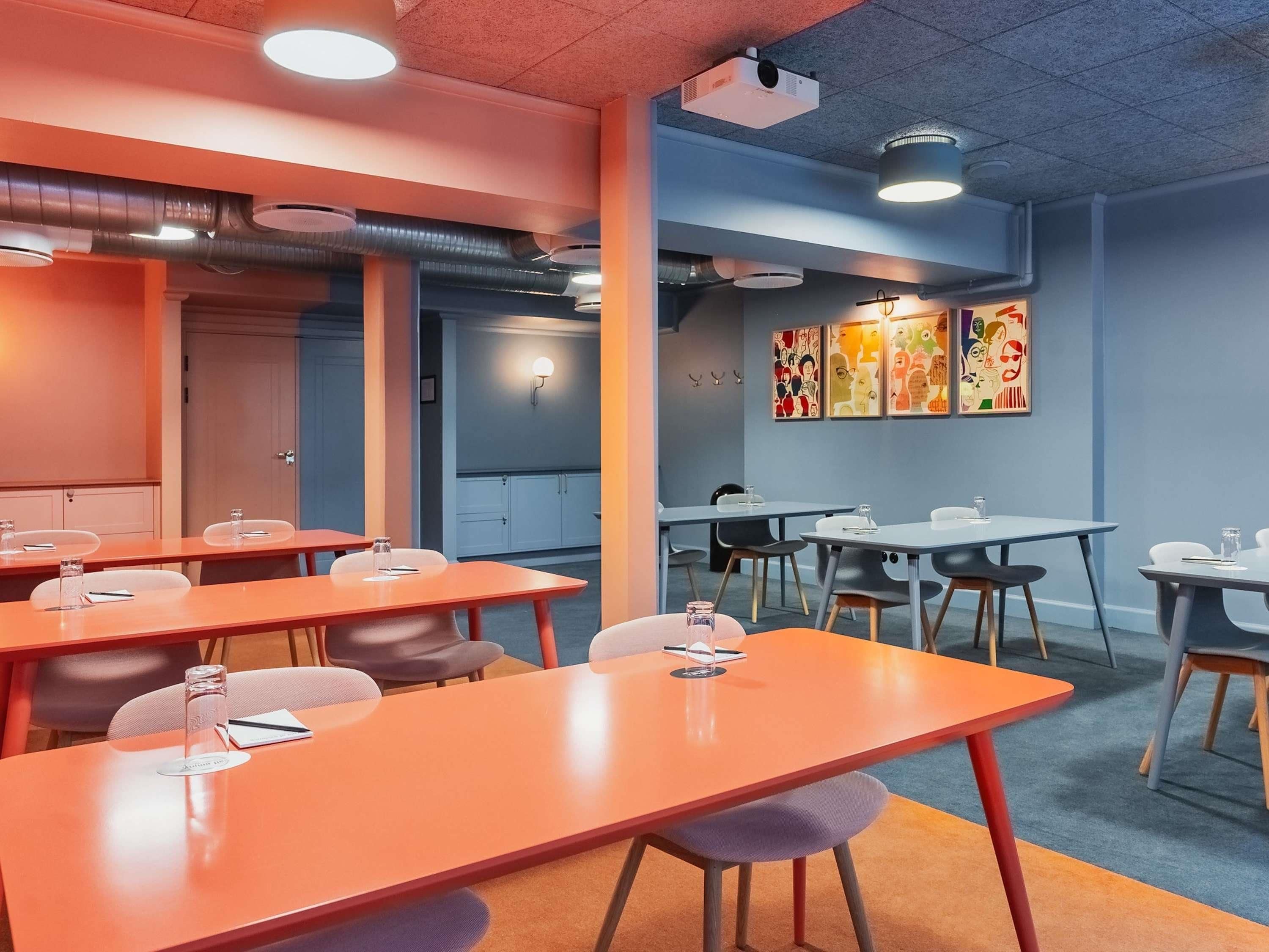 Sala de conferências com várias mesas e cadeiras, um projetor e obras de arte nas paredes.