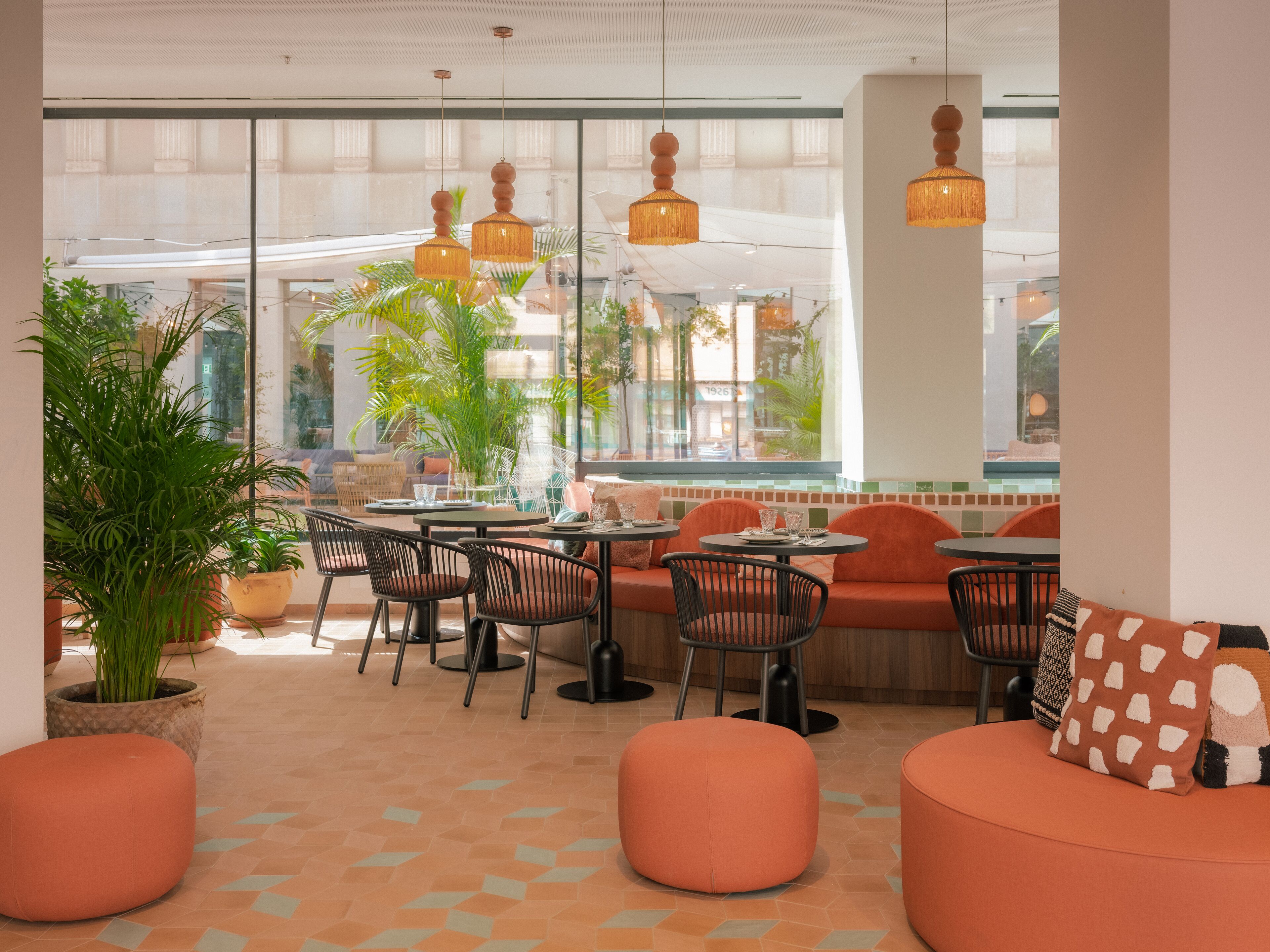 Uma área de estar interna moderna, com mesas redondas e assentos acolchoados em laranja, janelas amplas e luminárias pendentes.