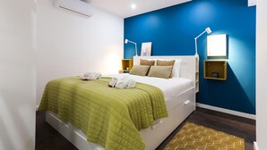 Quarto de hotel moderno com uma cama grande, colcha verde acolchoada, duas mesas de cabeceira com abajures e uma parede de destaque azul.