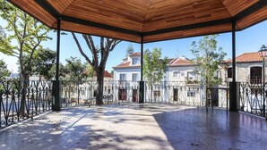 Uma área externa coberta, com teto de madeira, cercas de metal e vista para edifícios ao fundo.