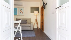 Um espaço de estar compacto com sofá, mesa pequena, luminária e TV fixada na parede.