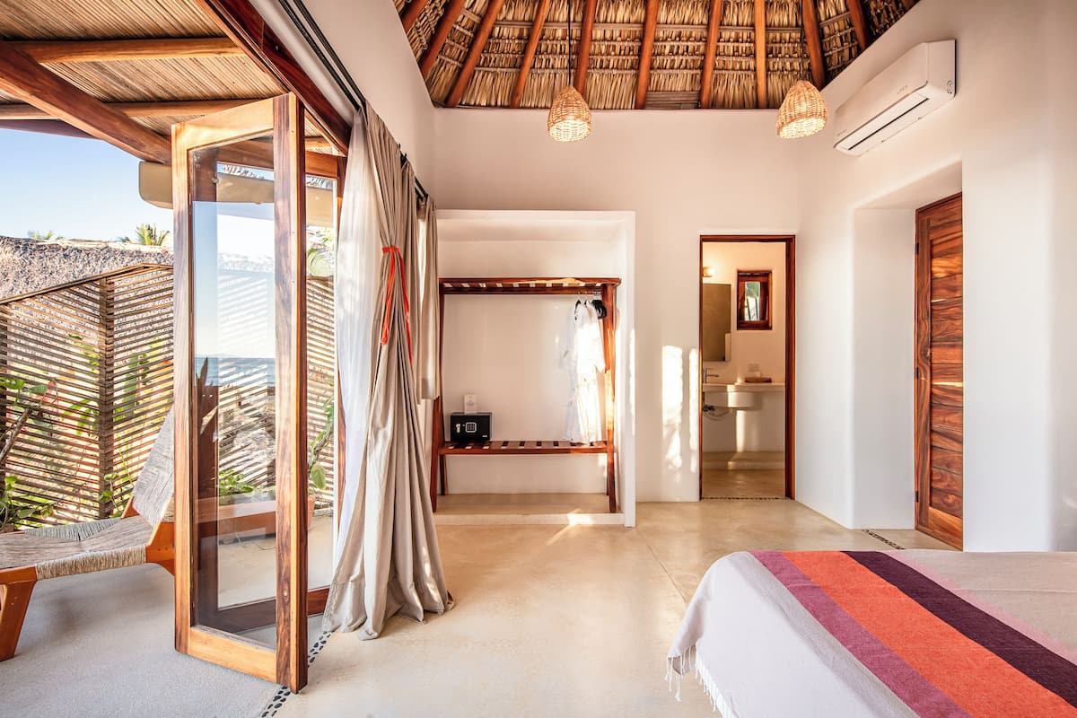 Quarto de hotel moderno com uma cama grande, um espaço de estar com um banco, uma porta de vidro que dá para uma área externa e um ventilador de teto.