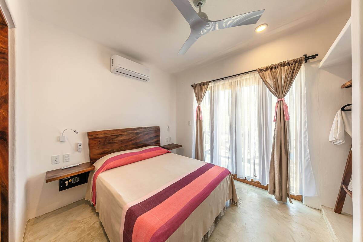 Quarto de hotel com cama, ventoinha no teto, ar condicionado, mesa de cabeceira e janela com cortinas.