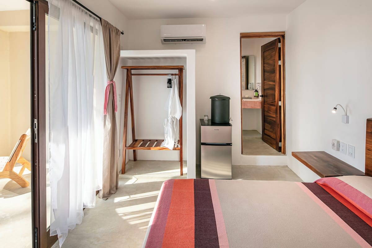 Quarto de hotel com cama, armário de madeira, pequena escrivaninha com espelho e zona de chuveiro.