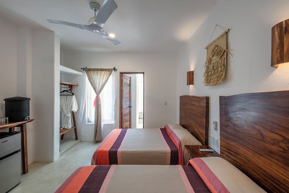 Quarto de hotel com duas camas, um ventilador de teto, cabeceira de madeira, luminária na parede e um guarda-roupa.