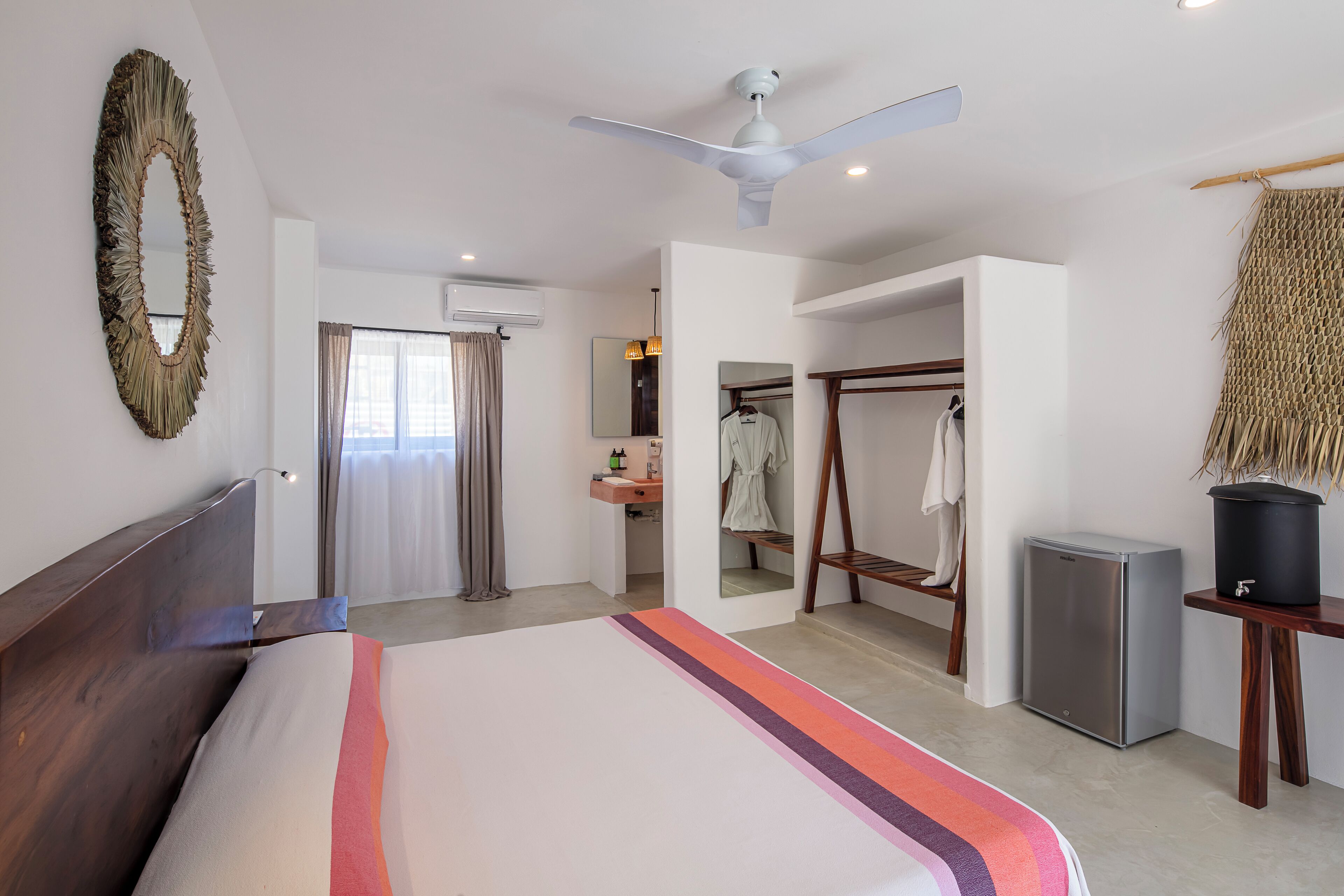 Quarto de hotel com cama, ventoinha no teto, guarda-roupa, mini-frigorífico e espelho na parede.
