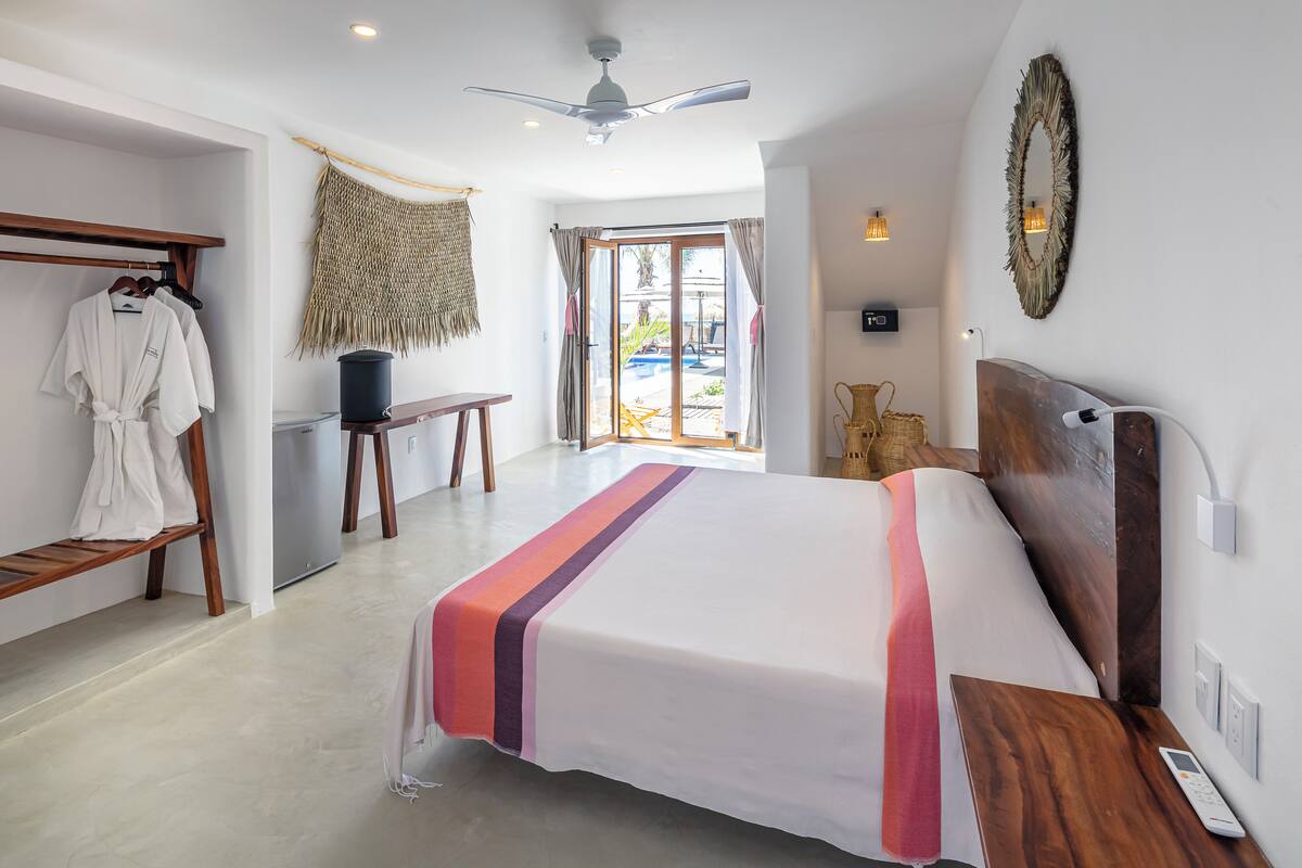 Quarto de hotel com cama, um banco de madeira, televisão, ventoinha no teto e vista para o exterior.