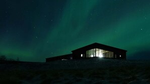Um edifício moderno de cor escura, com grandes janelas, localizado em uma colina sob um céu estrelado com aurora boreal visível.