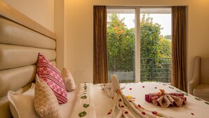 Cama bem arrumada com um arranjo floral, uma janela com cortinas e vista para o verde lá fora.