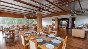 Uma ampla sala de jantar com várias mesas e cadeiras de madeira, janelas grandes e teto alto com vigas aparentes.