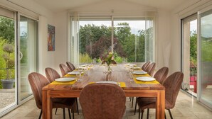 Uma sala de jantar com uma mesa grande posta para refeição, rodeada por cadeiras, grandes janelas e vista para o verde lá fora.