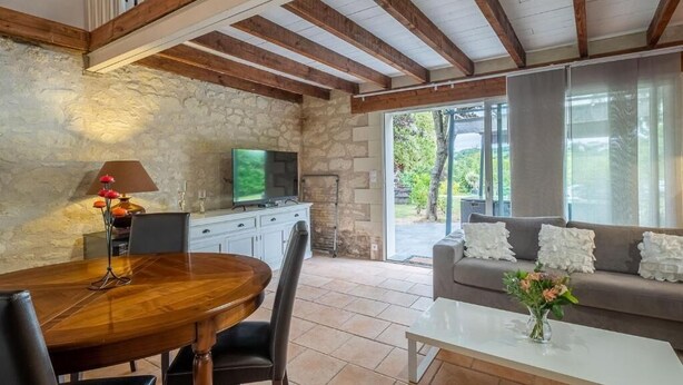 Uma sala de estar espaçosa com parede de pedra, vigas de madeira no teto, uma mesa de jantar, um sofá e uma televisão.