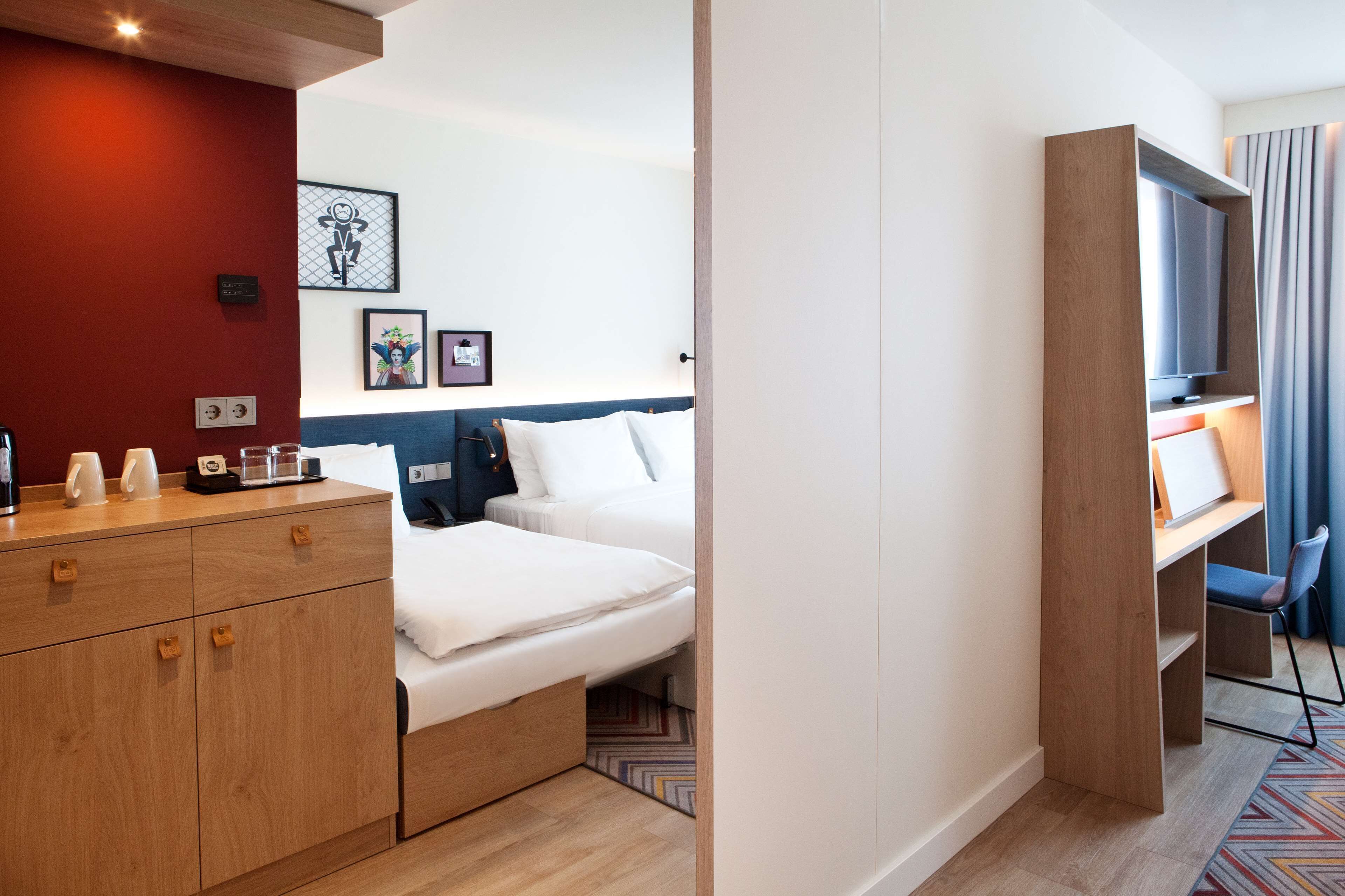 Quarto de hotel com cama, cómoda de madeira, escrivaninha com cadeira, televisão e obras de arte na parede.