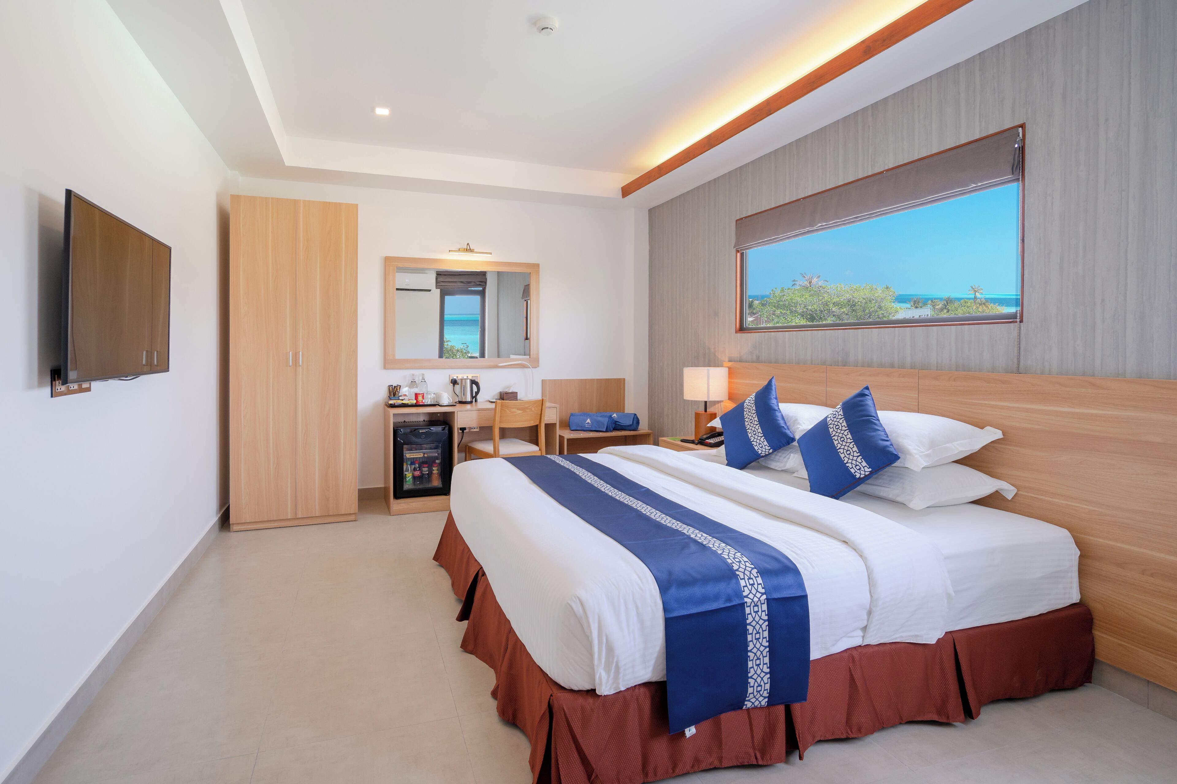 Quarto de hotel moderno com uma cama grande, uma escrivaninha com cadeira, um guarda-roupa e uma janela ampla com vista para o mar.