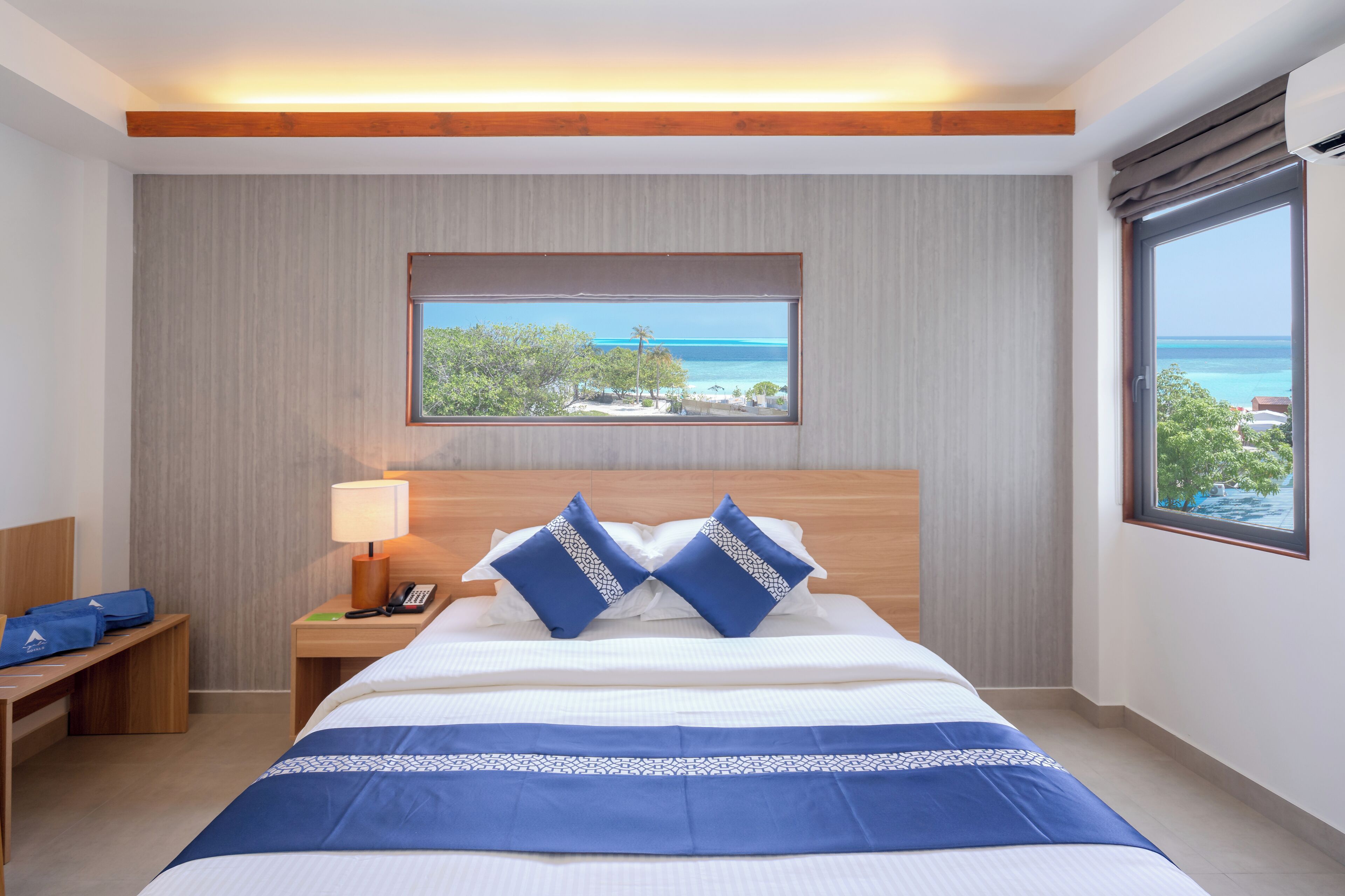 Quarto de hotel com uma cama grande, mesa de cabeceira com luminária, cabeceira de madeira, janela com vista para a praia e uma pequena mesa com uma cadeira.