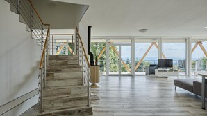 Interior moderno com escada de madeira, televisão de tela plana e janelas amplas com vista para o exterior.