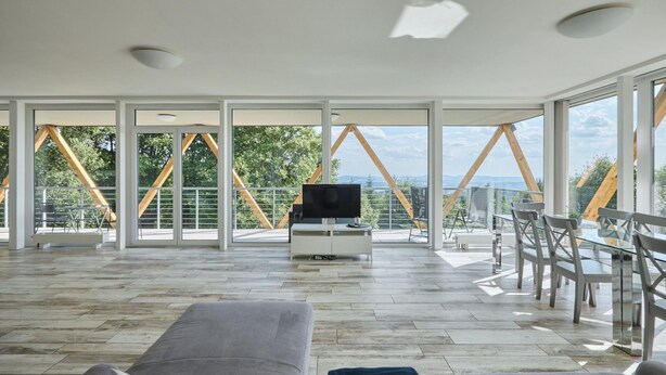Uma sala de estar ampla com um sofá moderno, uma televisão de tela plana e uma mesa de jantar com cadeiras. Janelas grandes oferecem vista para o exterior.
