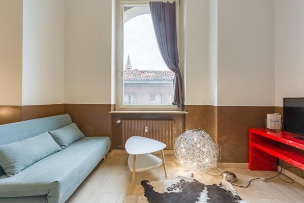 Uma sala de estar moderna com um sofá, uma poltrona branca, uma luminária decorativa e uma televisão sobre um suporte vermelho.