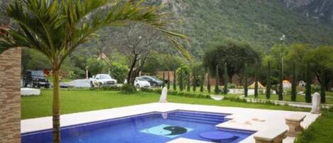Piscina com montanhas ao fundo, cercada por um gramado bem cuidado e algumas árvores.