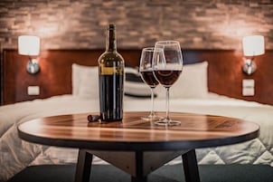 Mesa de madeira redonda com uma garrafa e duas taças de vinho sobre uma cama, com um muro de tijolos ao fundo.