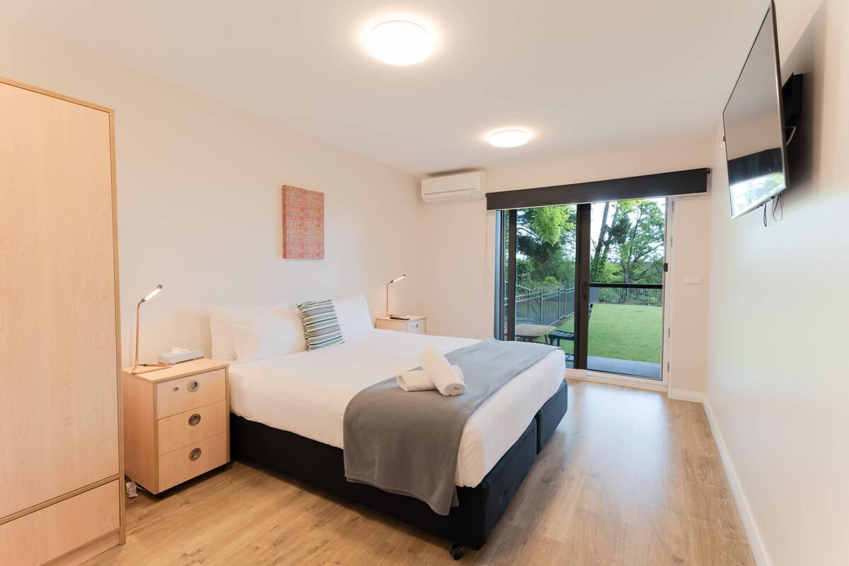 Quarto moderno com cama, mesas de cabeceira, televisão e porta de vidro deslizante para uma varanda.