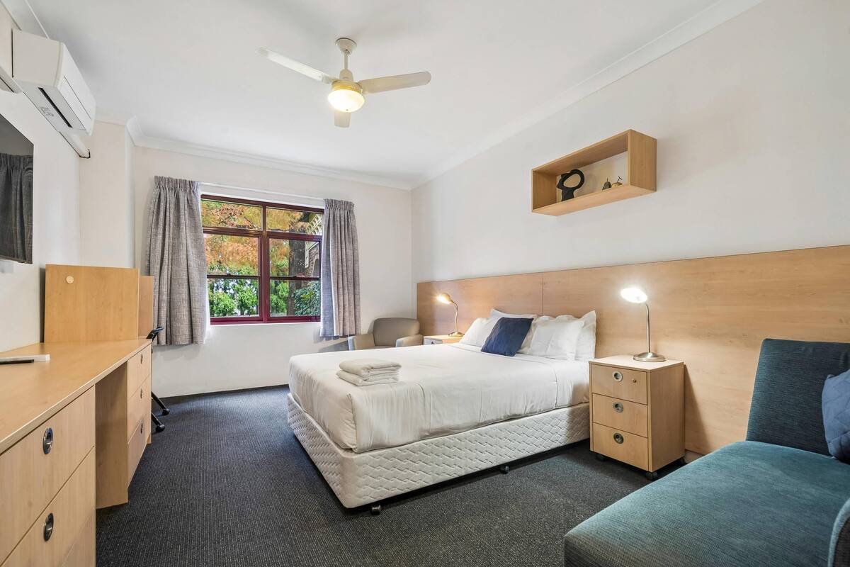 Quarto de hotel com cama, escrivaninha, cadeira, mesa de cabeceira e janela com cortinas.