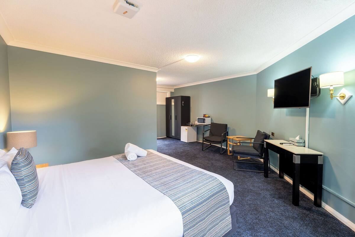 Quarto de hotel com cama, escrivaninha, cadeira, televisão e abajur.