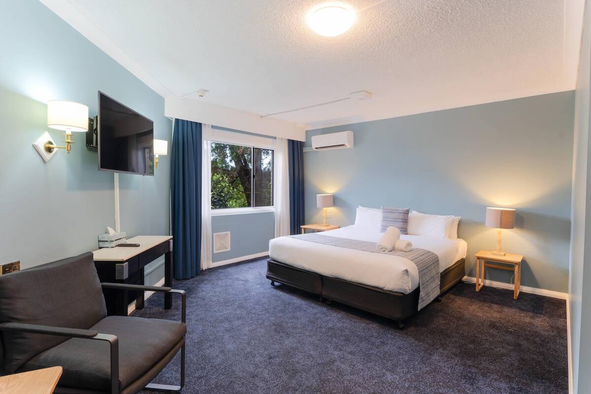 Quarto de hotel com cama, escrivaninha, cadeira, televisão e janela com cortinas.