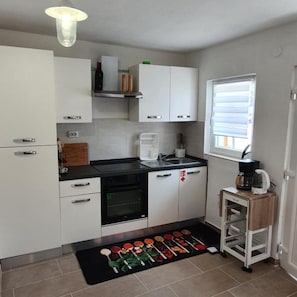 Uma cozinha compacta com armários brancos, bancada preta, forno embutido, cafeteira e uma janela com persianas.