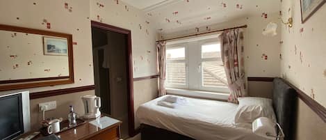 Quarto de hotel com cama, escrivaninha com televisão, cadeira, mesa de cabeceira com abajur e janela com cortinas.