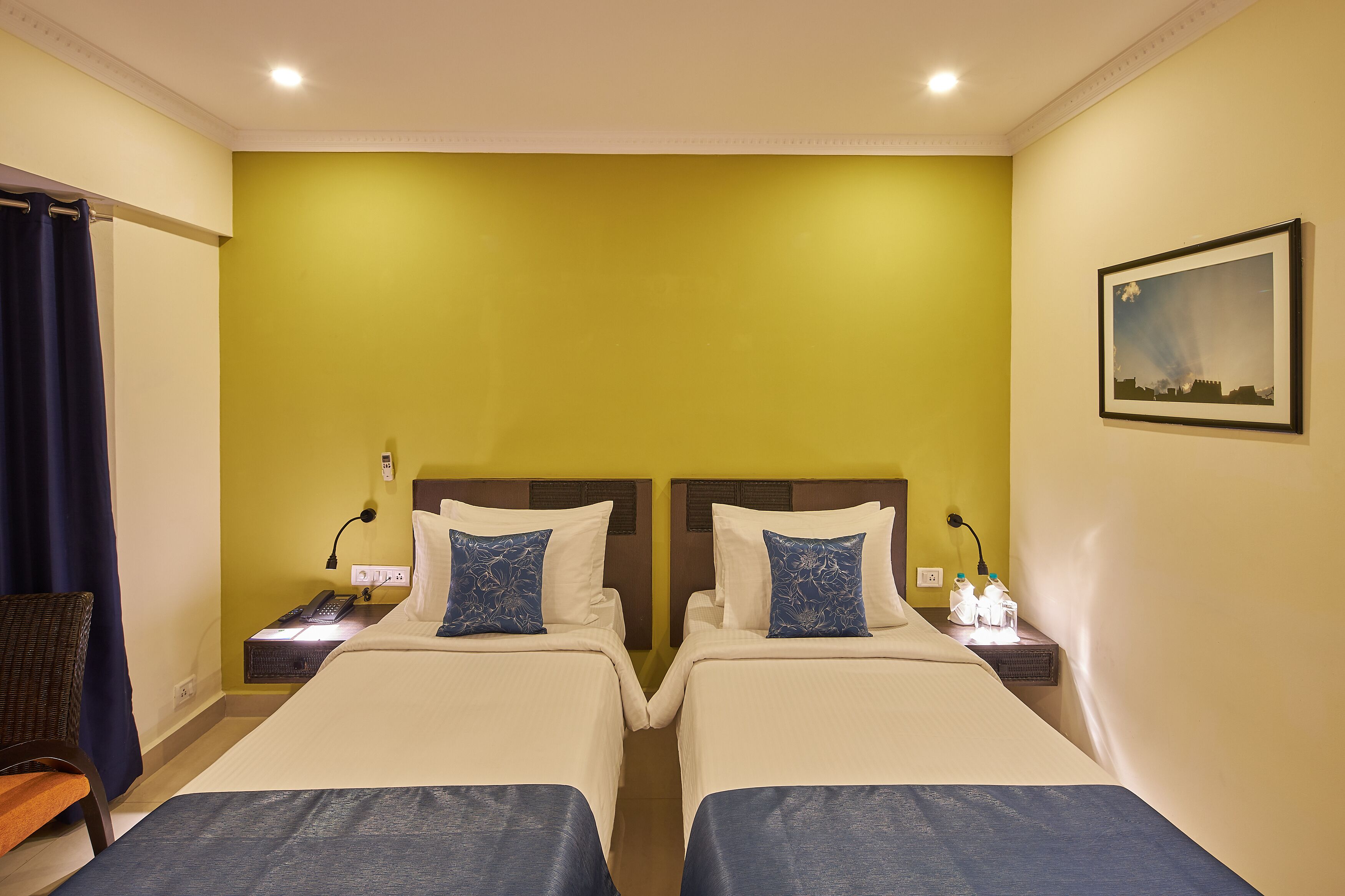 Quarto de hotel com duas camas, uma mesa de cabeceira com telefone e abajur, uma cadeira e um quadro na parede.