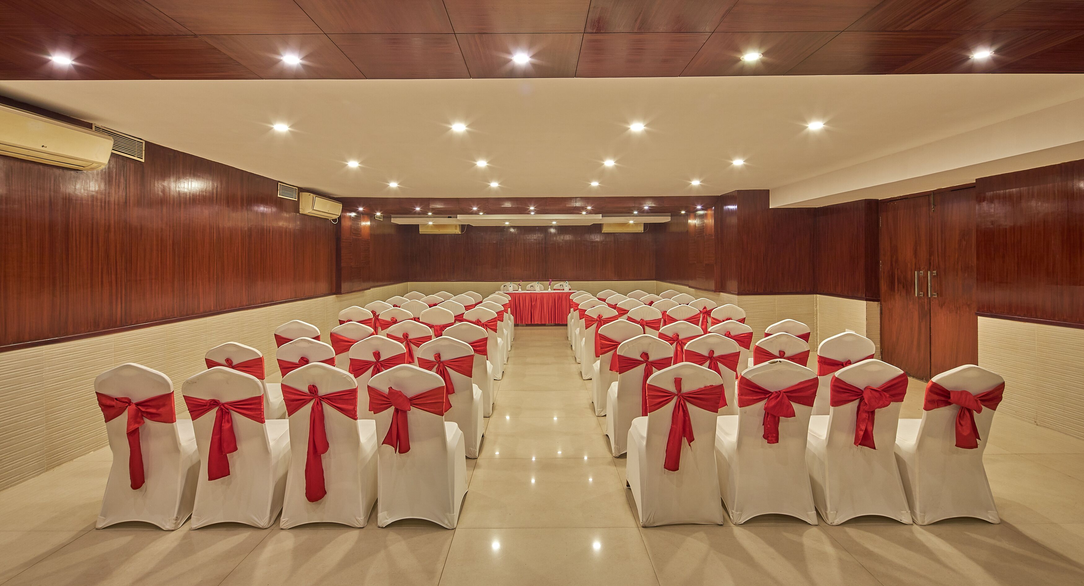 Uma sala de conferência com fileiras de cadeiras brancas com fitas vermelhas, painéis de madeira nas paredes e iluminação embutida.