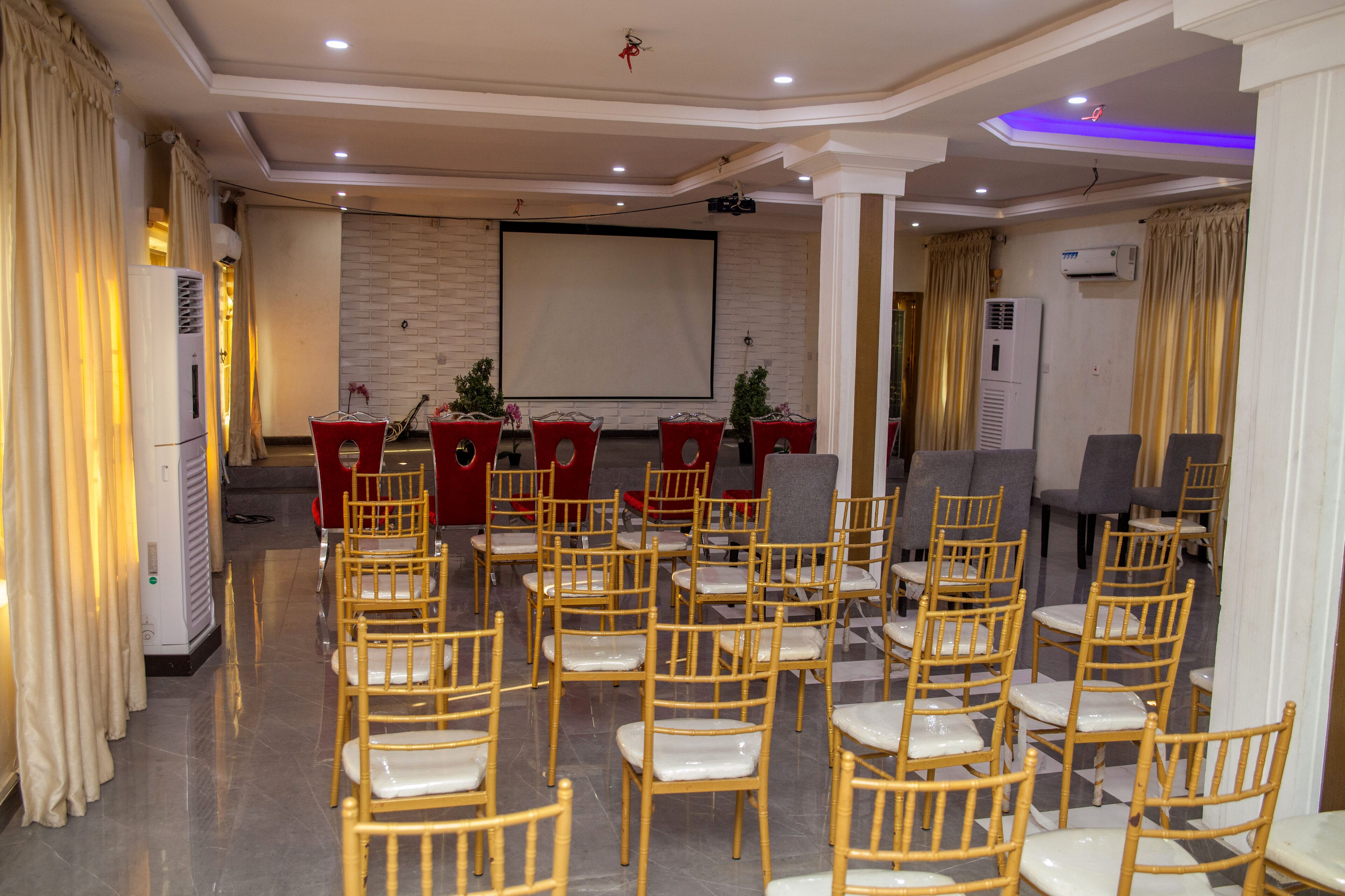 Uma sala de conferências com fileiras de cadeiras e mesas de madeira, uma tela de projeção e aparelhos de ar condicionado.