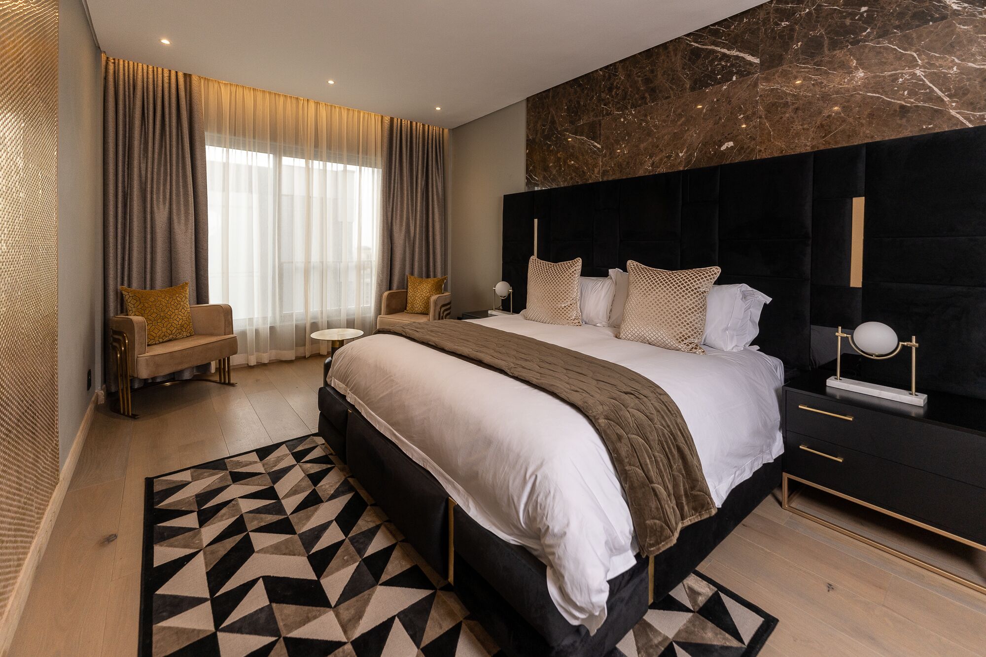 Quarto moderno com cama grande, cabeceira com textura que imita mármore, mesa de cabeceira com abajur e tapete com padrão geométrico.